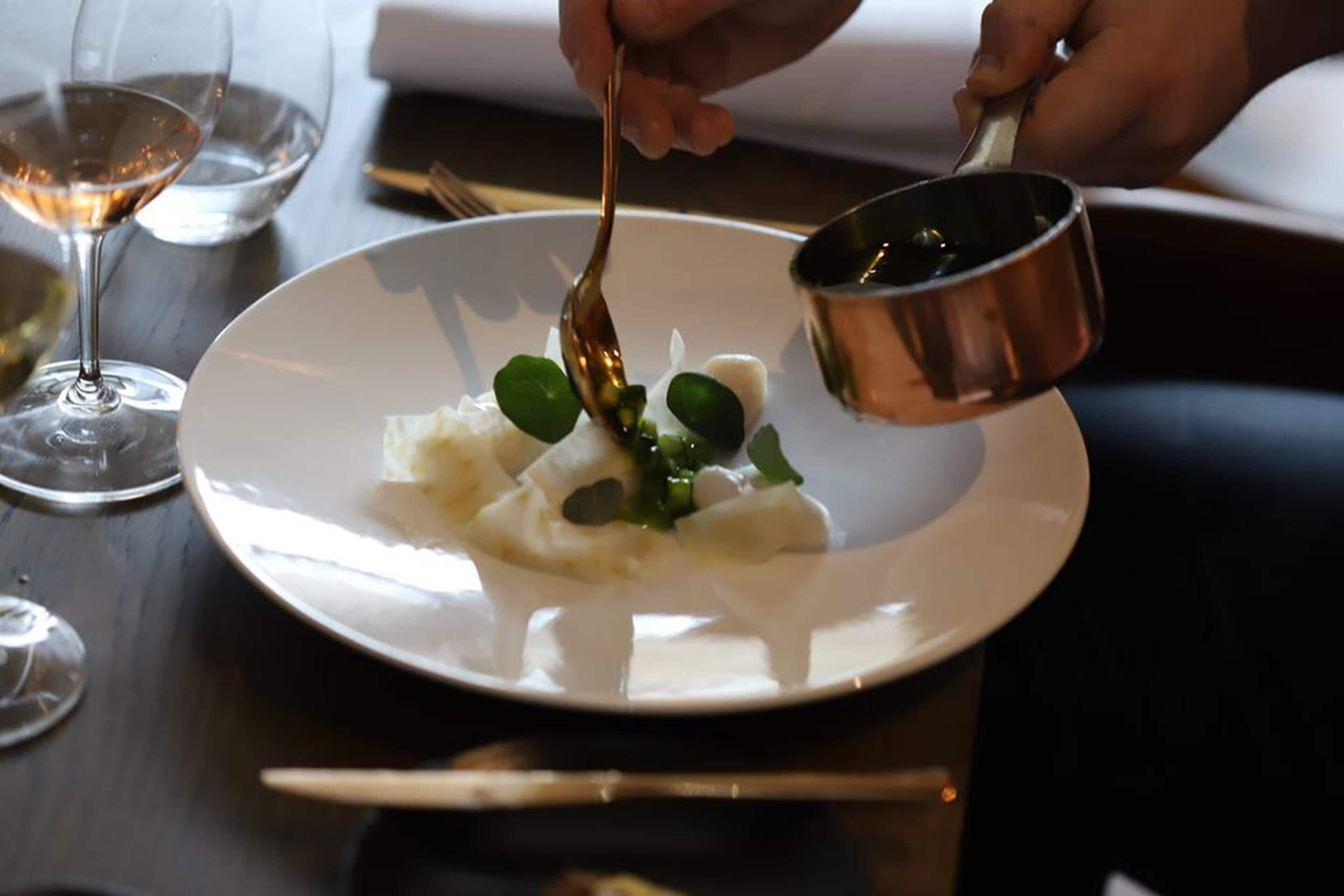 Uma pessoa despejando um líquido de uma panela de cobre sobre um prato branco com comida e folhas verdes.