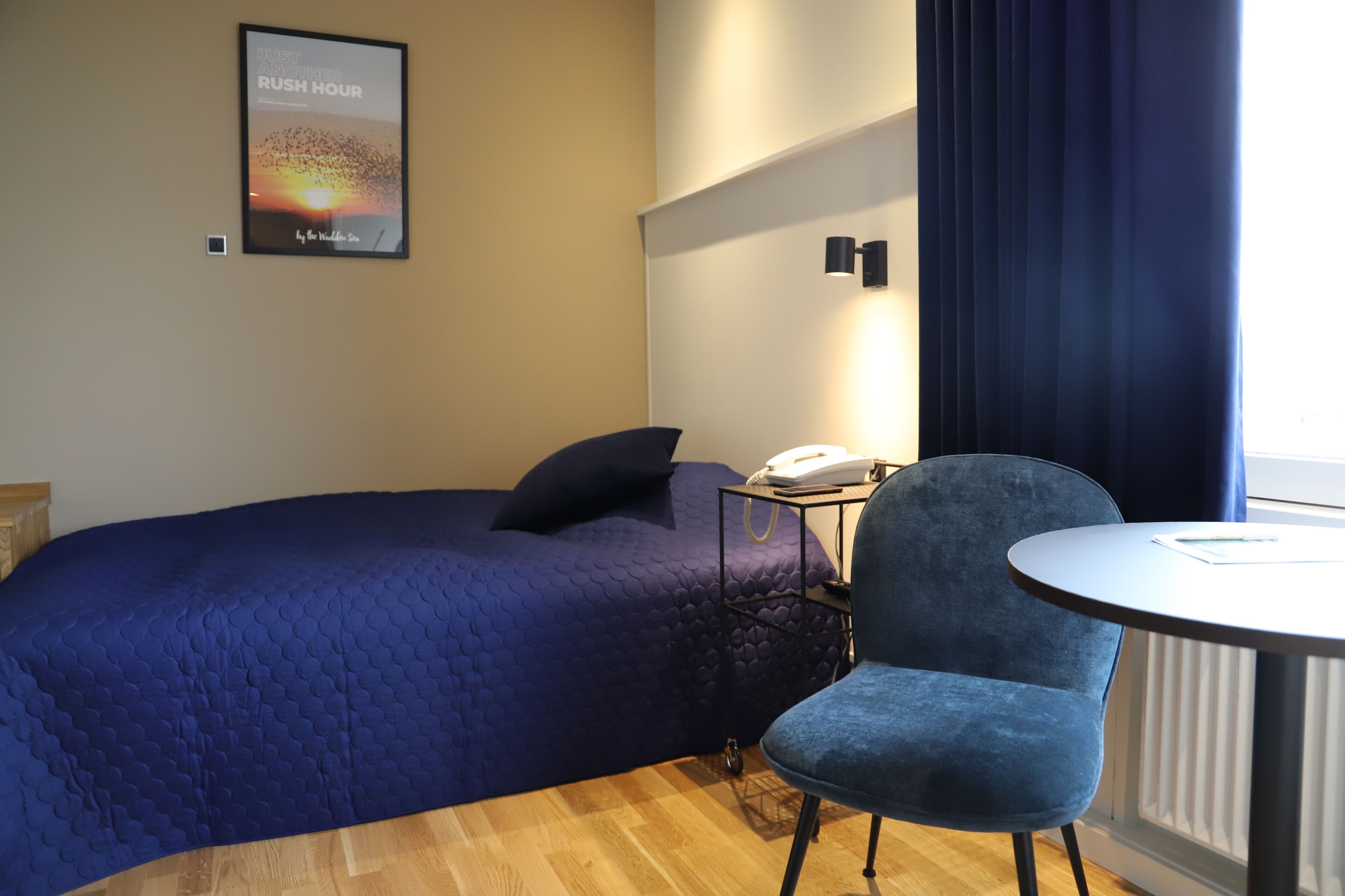 Quarto de hotel com uma cama grande, uma cadeira, uma mesinha, uma mesaninha de cabeceira, um abajur e uma janela com cortinas.