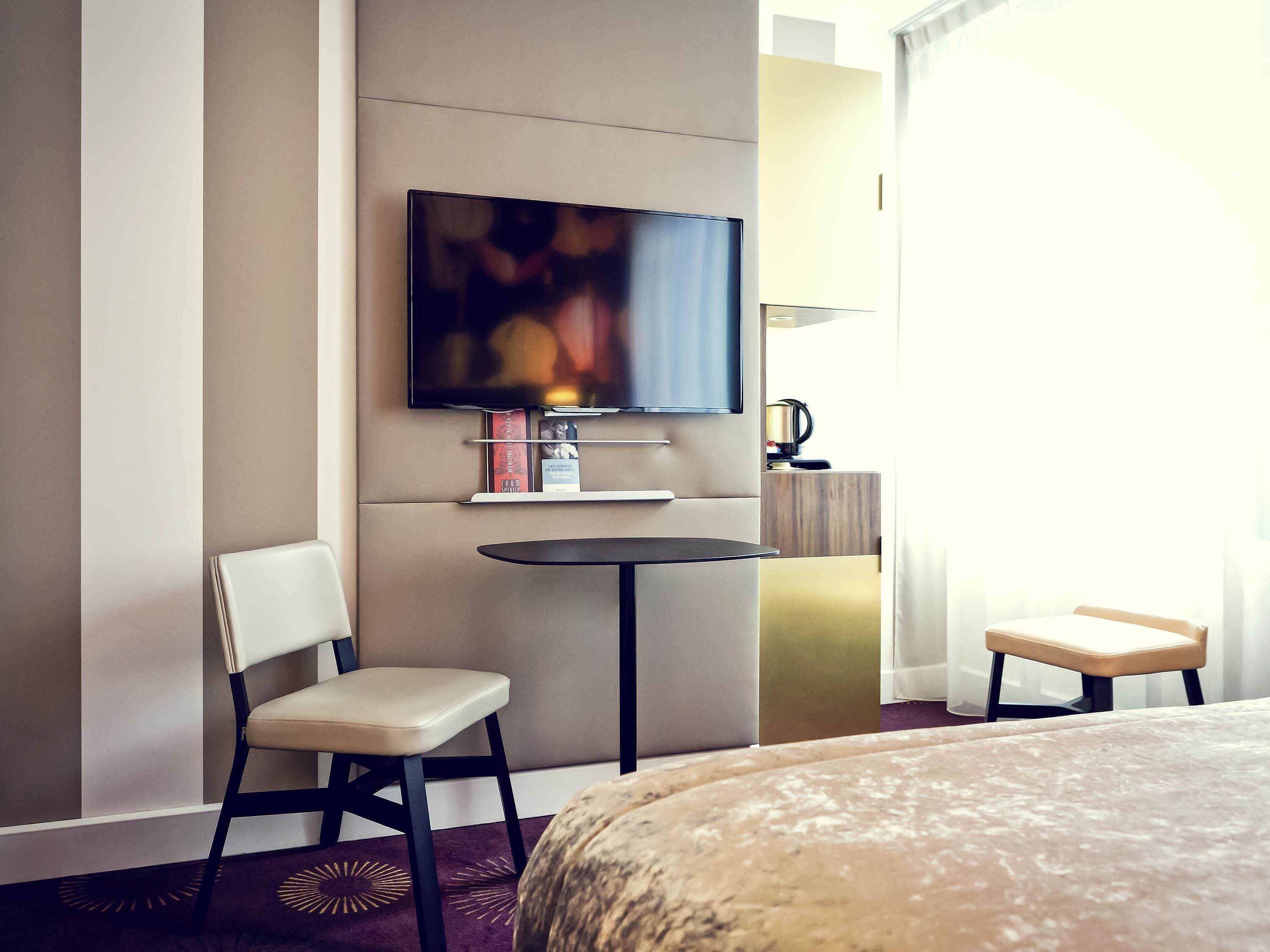 Quarto de hotel com cama, televisão na parede, mesa pequena, duas cadeiras e máquina de café.