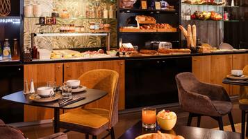 Um balcão de café da manhã com diversas opções de alimentos, incluindo doces e frutas. Há áreas de estar com cadeiras e mesas, além de um balcão com uma máquina de café.