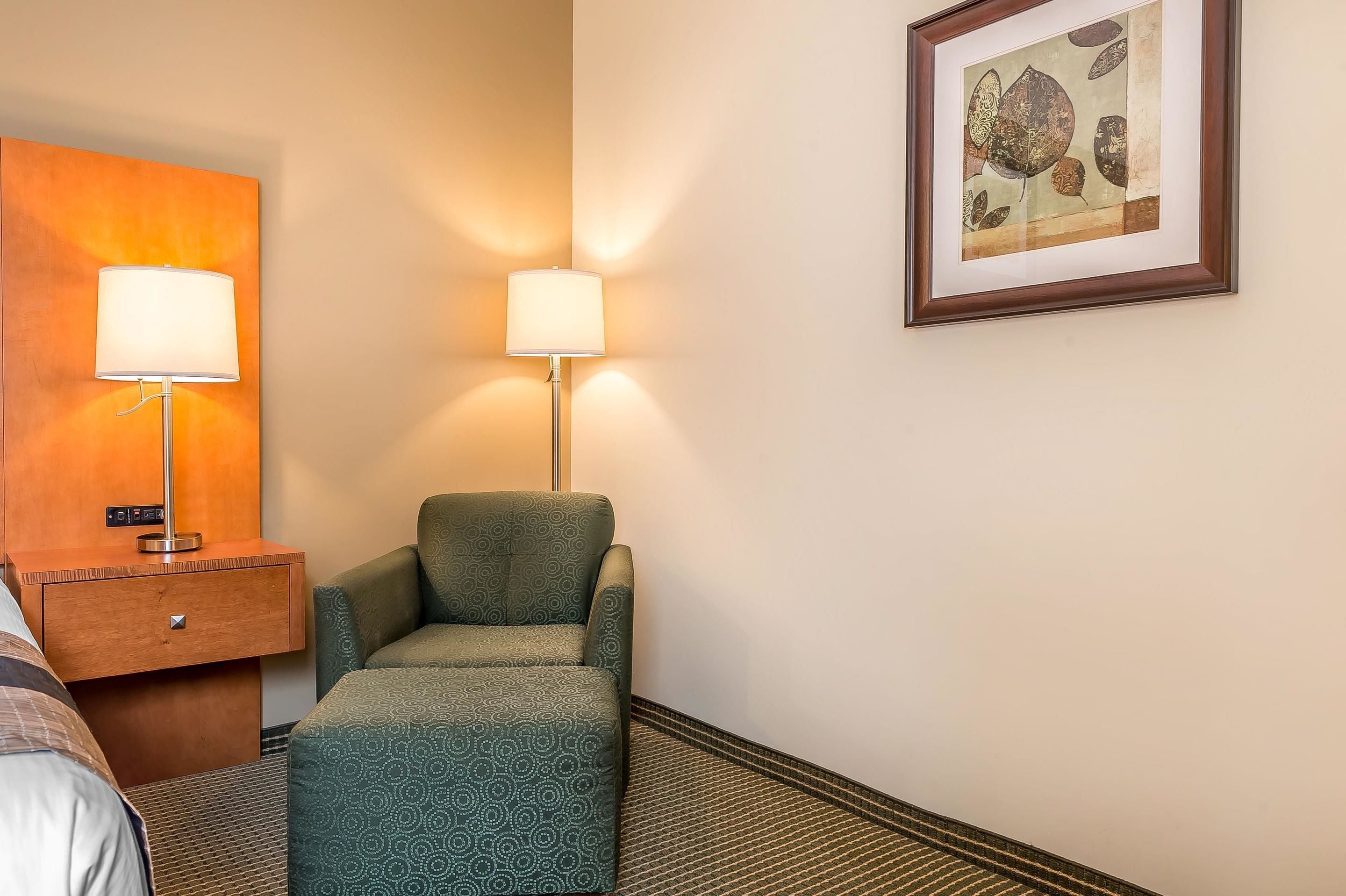 Quarto de hotel com cama, poltrona verde, mesa de cabeceira com abajur e uma obra de arte emoldurada na parede.