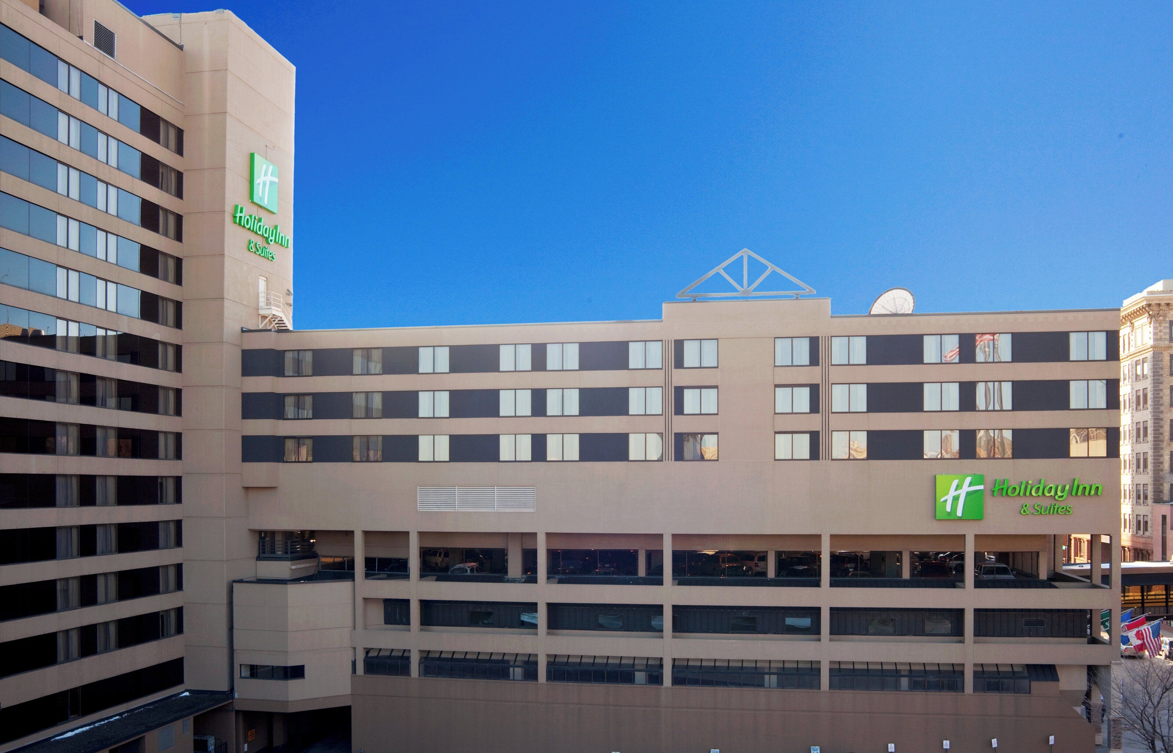 Edifício de vários andares do Holiday Inn & Suites com um logotipo 'H' em destaque, grandes janelas e uma garagem subterrânea.