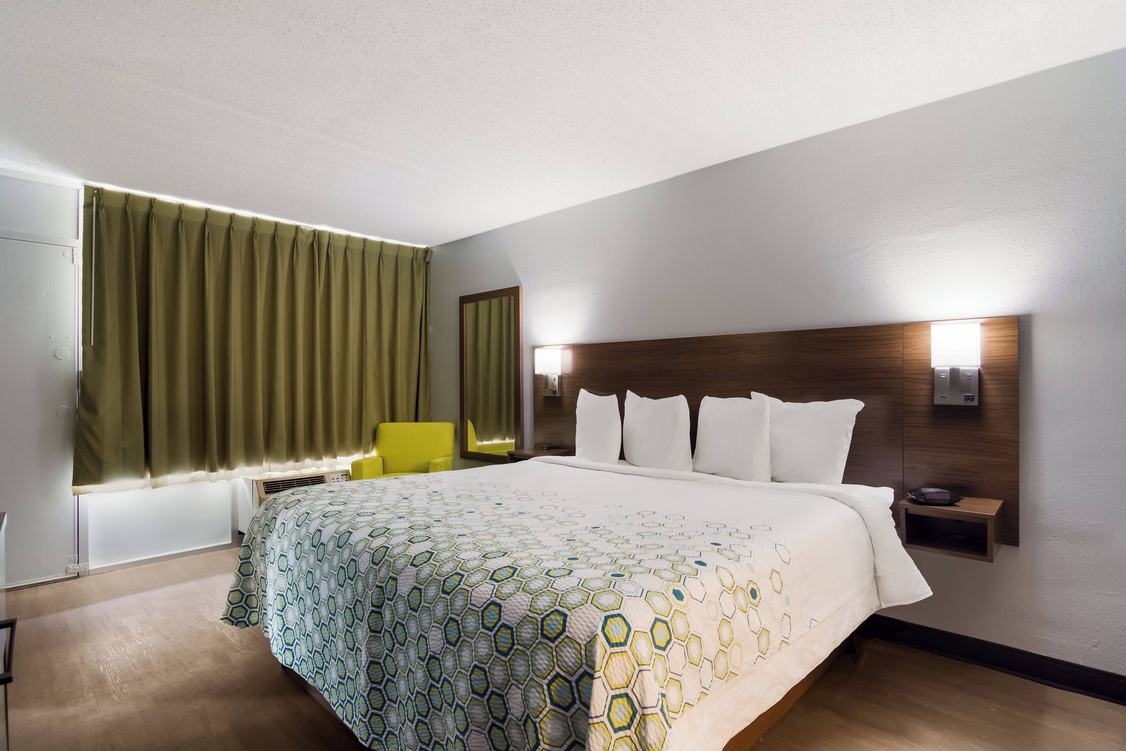Quarto de hotel com uma cama grande, mesa de cabeceira, espelho e janela com cortinas.