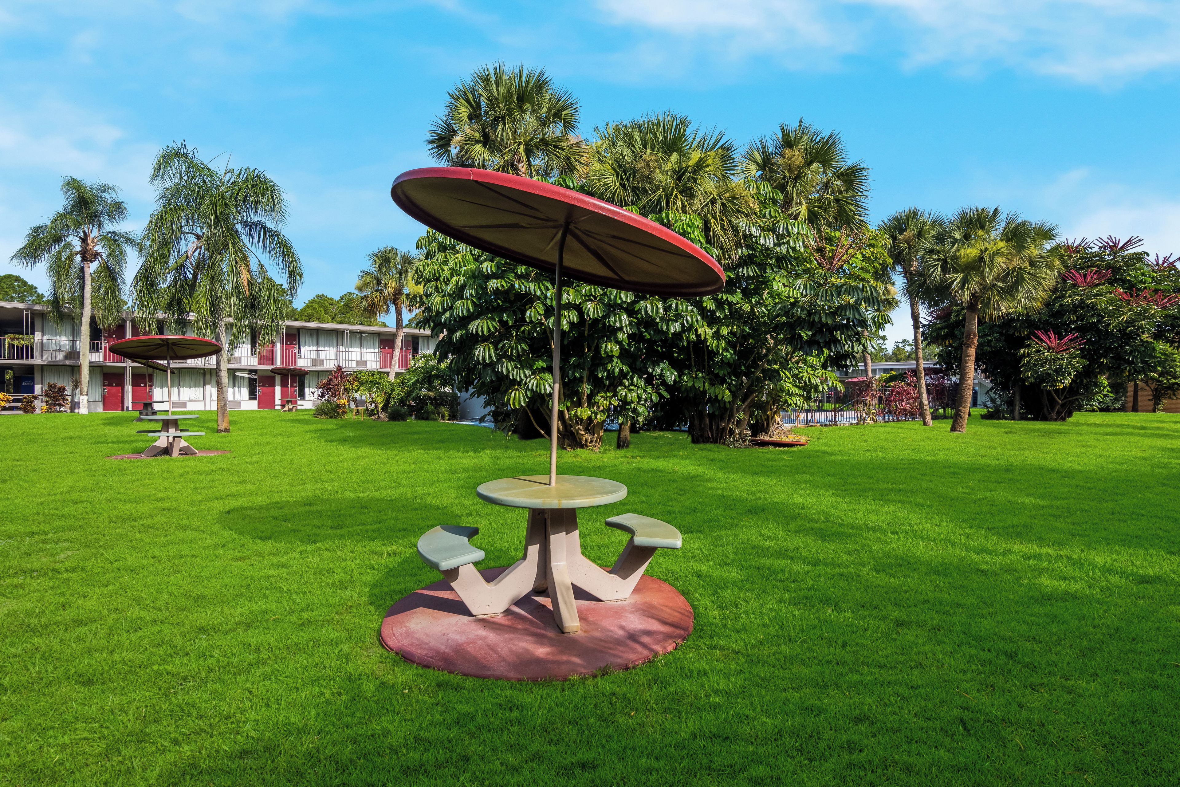Uma área gramada bem cuidada, com uma mesa e guarda-sol no centro, rodeada por vegetação exuberante e um edifício com várias janelas ao fundo.