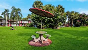 Uma área gramada bem cuidada, com uma mesa e guarda-sol no centro, rodeada por vegetação exuberante e um edifício com várias janelas ao fundo.