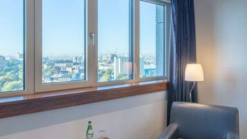Quarto de hotel com uma janela grande, uma mesa de madeira com garrafa e copo, uma poltrona azul e um abajur.