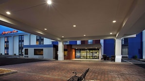 Entrada de um hotel Hampton Inn com área externa coberta, calçada de tijolos e um banco.