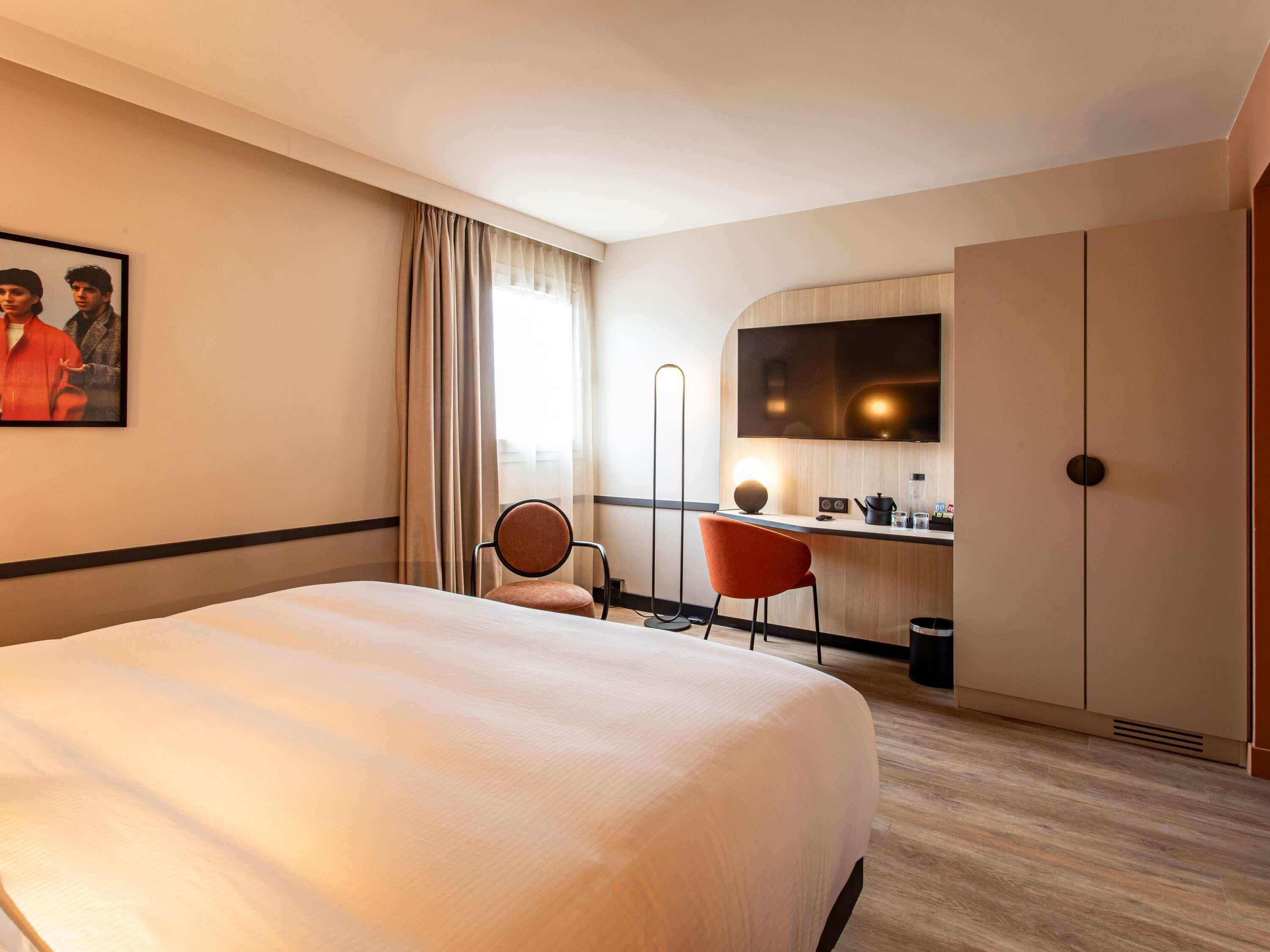 Quarto de hotel com uma cama grande, uma escrivaninha com cadeira, uma televisão e um guarda-roupa.