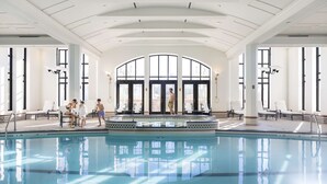 Uma grande piscina coberta com paredes de vidro transparente, várias áreas de descanso e uma escadaria central.