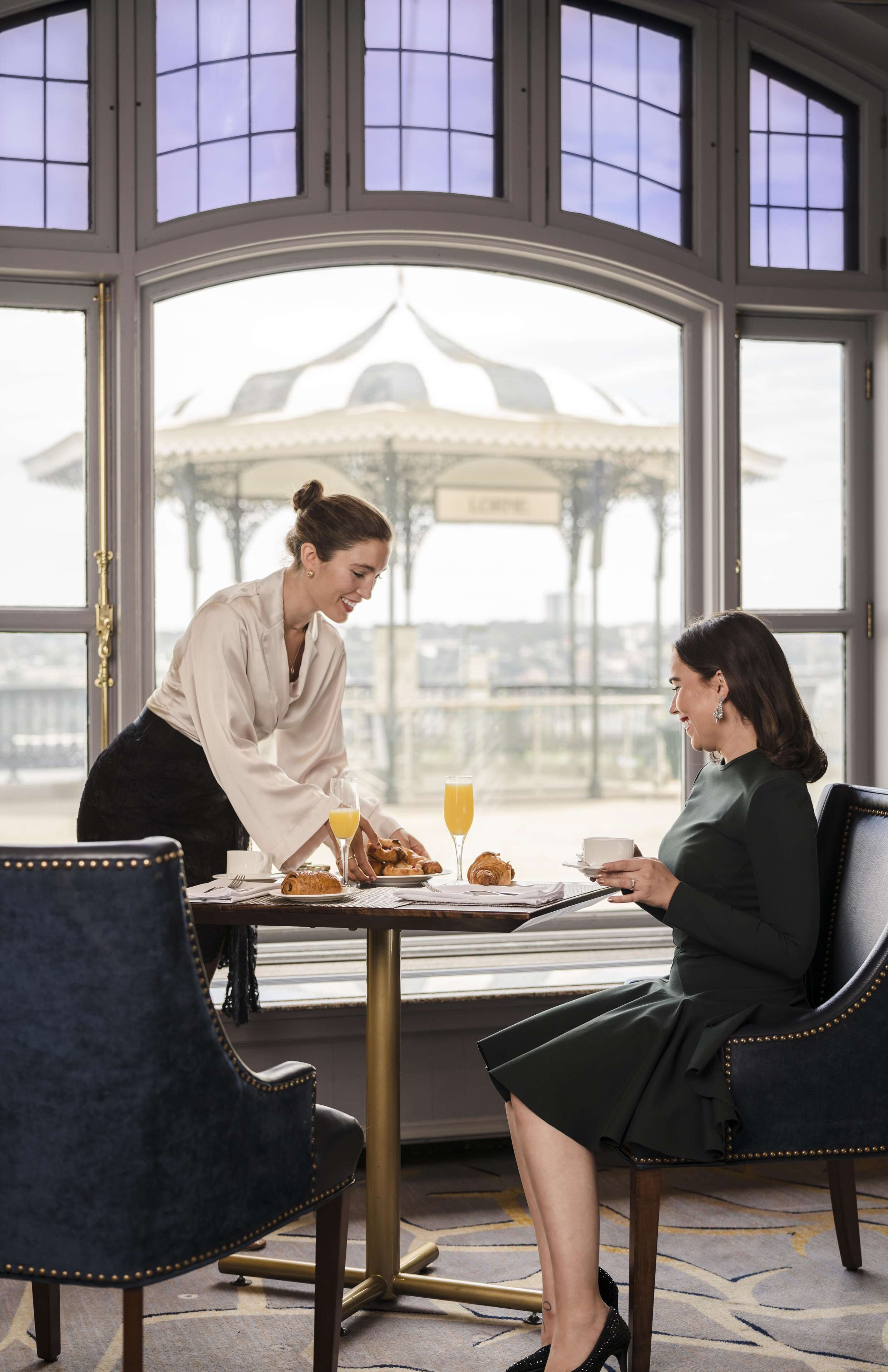 Uma área de refeições com uma mesa posta para dois, um garçom servindo comida e vista para um edifício através de grandes janelas.