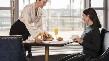 Uma área de refeições com uma mesa posta para dois, um garçom servindo comida e vista para um edifício através de grandes janelas.