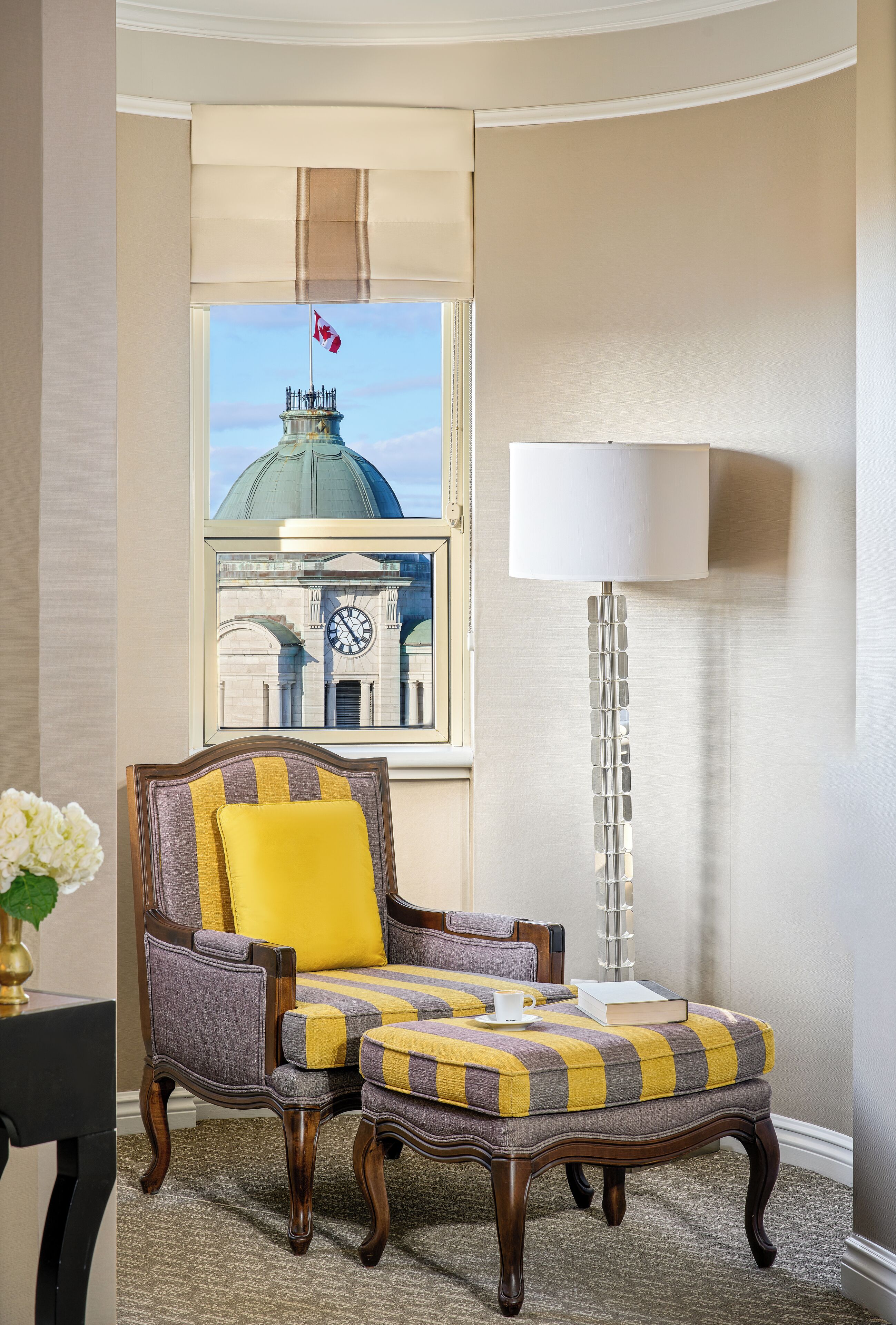 Quarto de canto com vista para um edifício através de uma janela, uma luminária, uma cadeira com almofada e uma pequena mesa com um vaso.