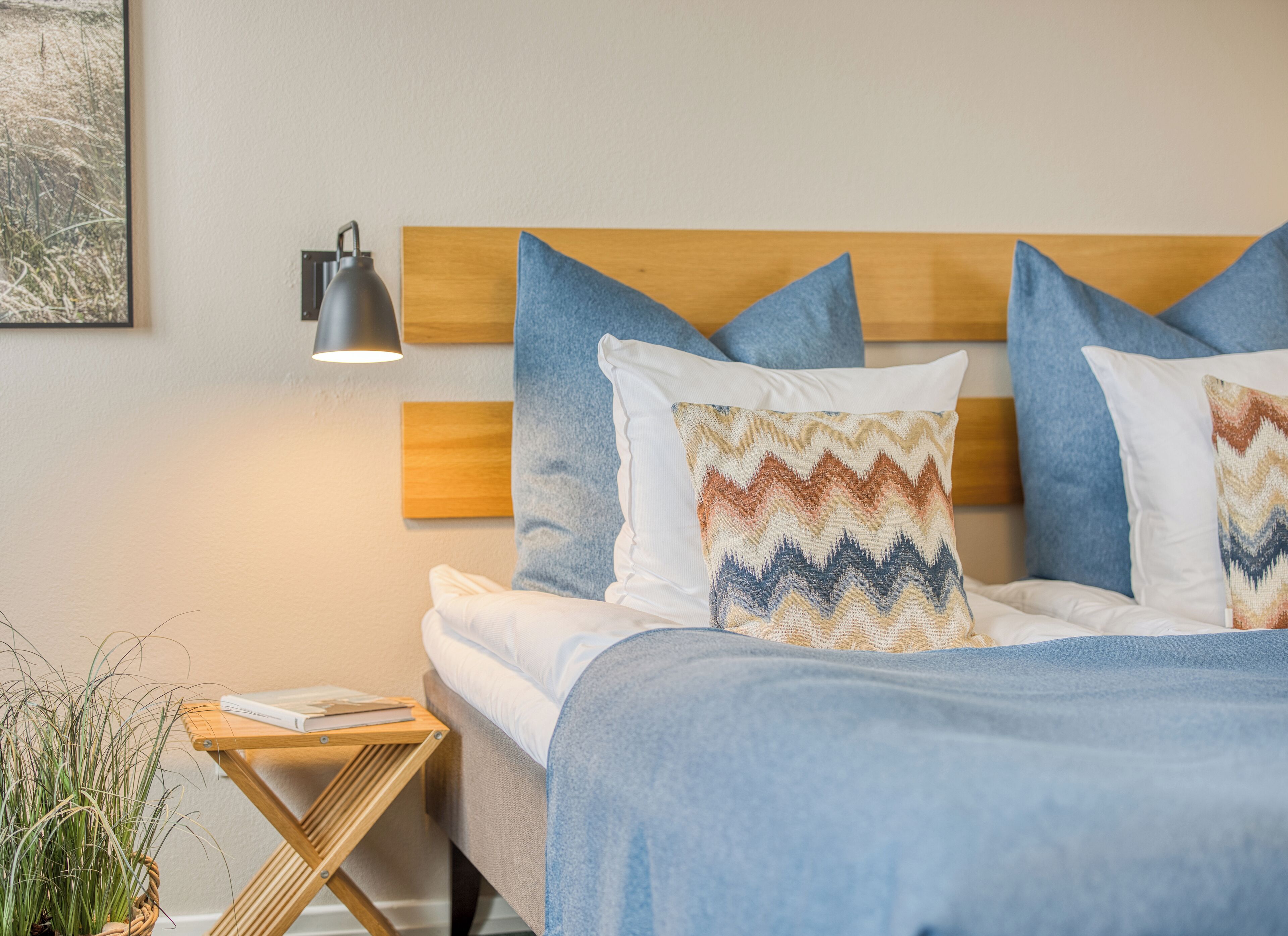 Cama bem arrumada com manta azul e almofadas estampadas, mesa de cabeceira com abajur e uma obra de arte na parede.