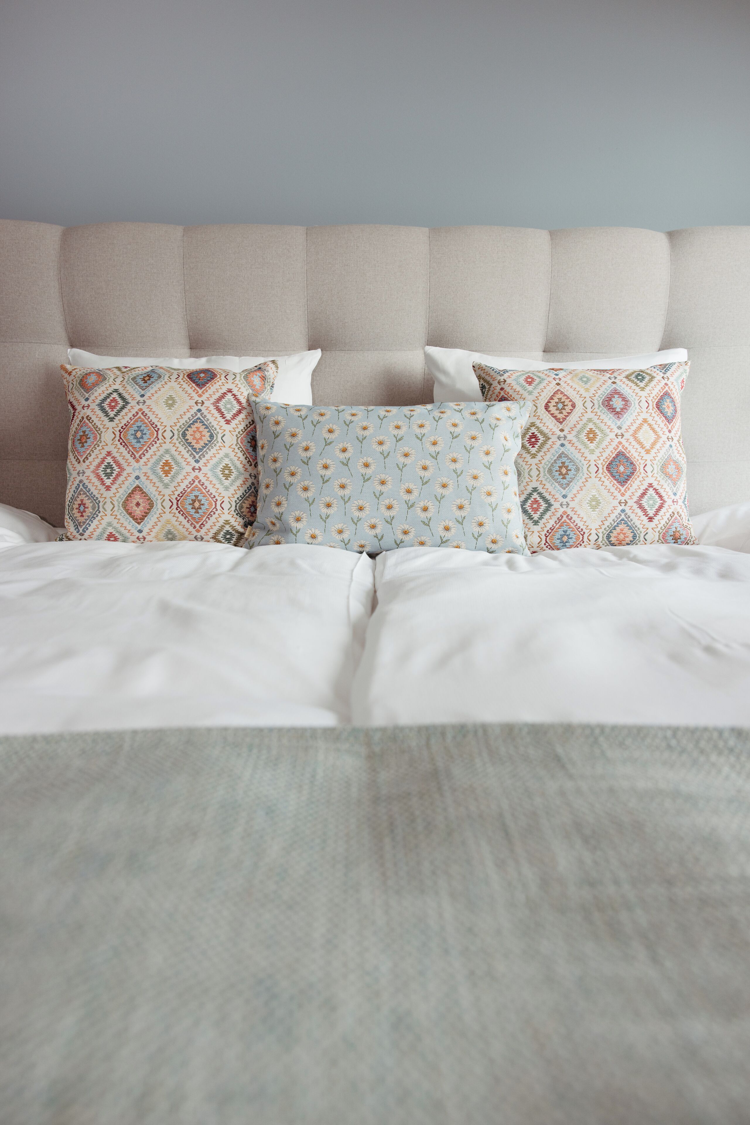 Cama com lençóis brancos, cabeceira estofada em bege e almofadas estampadas.