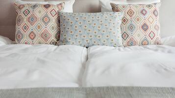 Cama com lençóis brancos, cabeceira estofada em bege e almofadas estampadas.