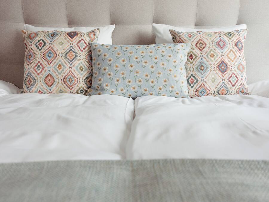 Cama com lençóis brancos, cabeceira estofada em bege e almofadas estampadas.