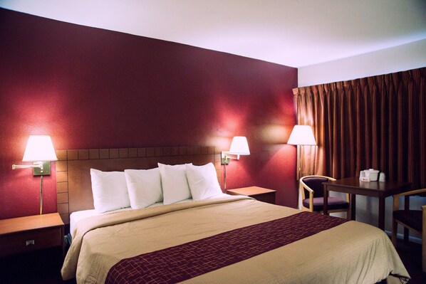 Quarto de hotel com uma cama grande, duas luminárias de cabeceira, uma mesinha de madeira, uma pequena mesa com uma cadeira e uma cortina na janela.