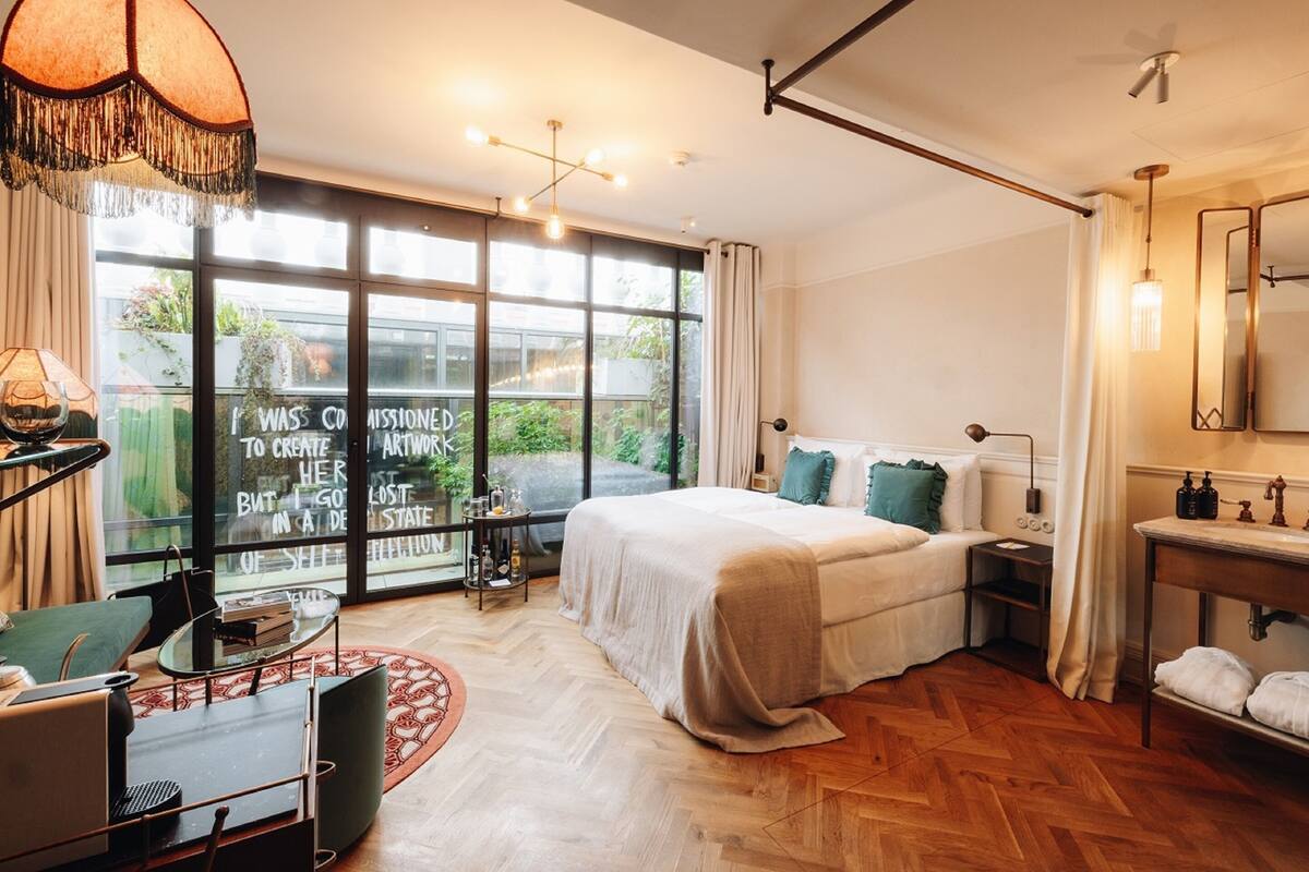 Quarto de hotel moderno com uma cama grande, uma escrivaninha, uma cadeira, uma mesa redonda, uma janela ampla e um banheiro com espelho e pia.