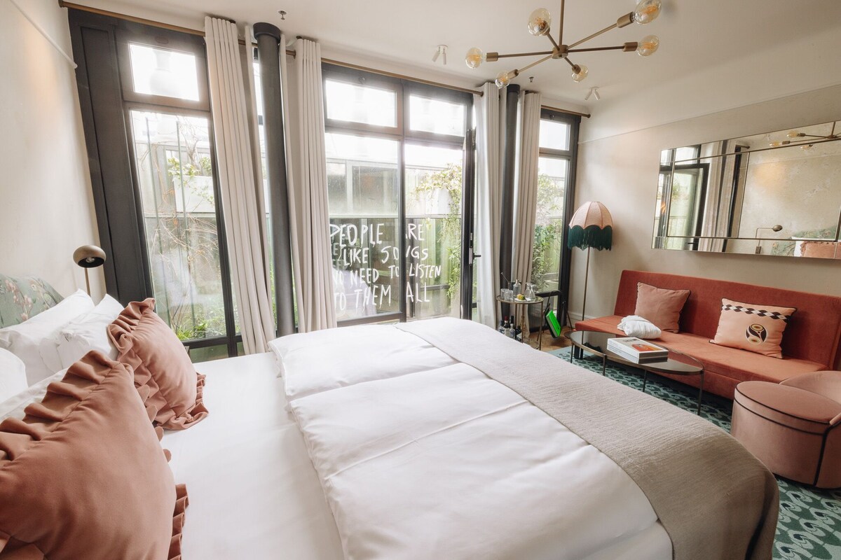 Quarto de hotel com uma cama grande, um sofá, uma mesinha, um espelho e vista para o exterior.