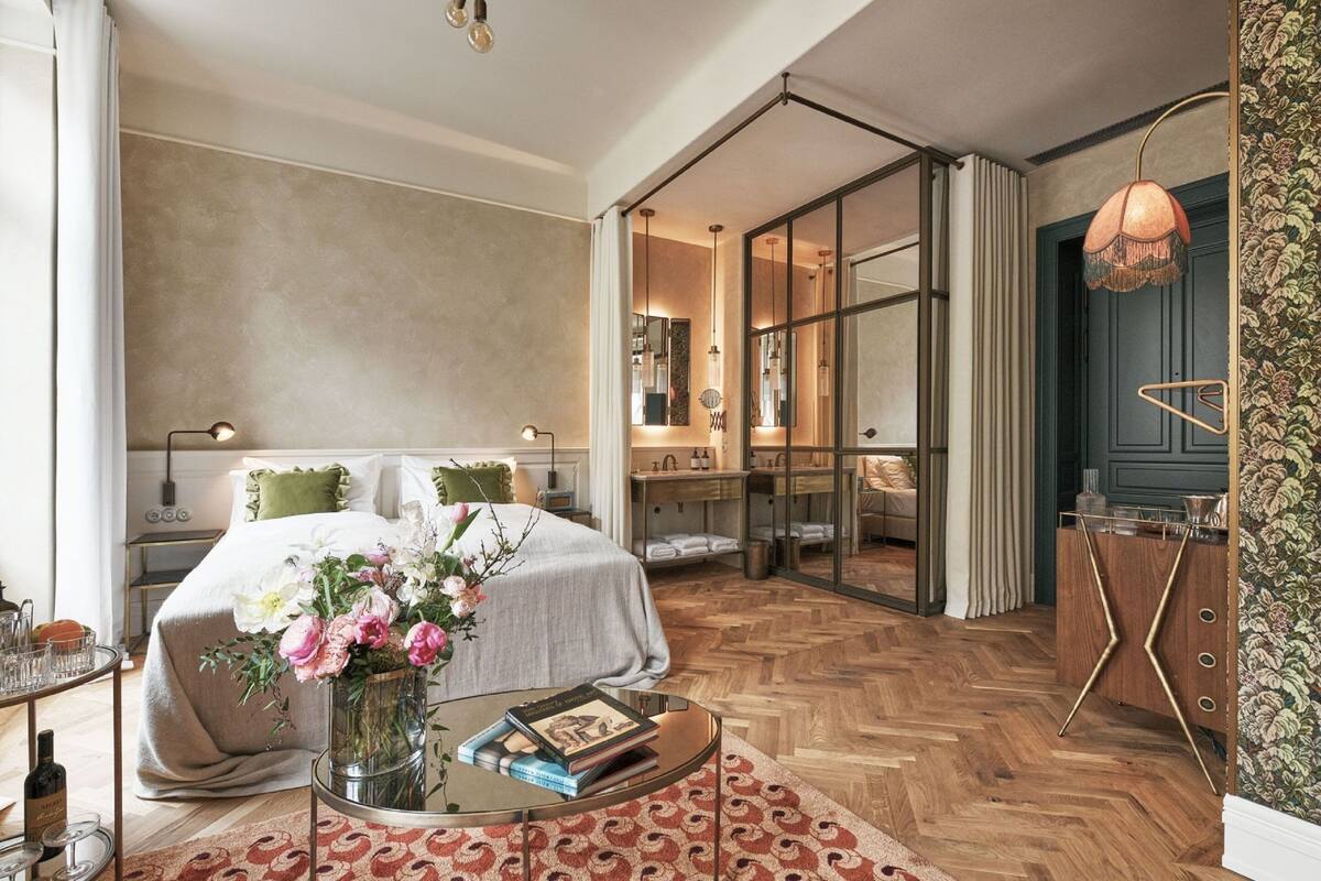 Quarto de hotel moderno com uma cama grande, casa de banho com paredes de vidro, soalho de madeira em padrão herringbone e uma pequena mesa redonda com um vaso de flores.
