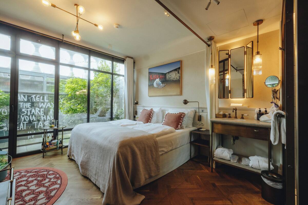 Quarto de hotel moderno com uma cama grande, piso de madeira, vista através de janelas amplas, um quadro na parede e um banheiro com espelho e porta-toalhas.