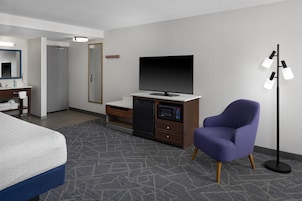 Quarto de hotel com cama, escrivaninha com televisão de tela plana, micro-ondas, cadeira e abajur.