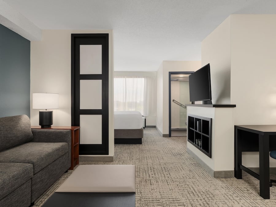 Quarto de hotel com sofá, cama, escrivaninha, televisão e banheiro visível por uma porta aberta.