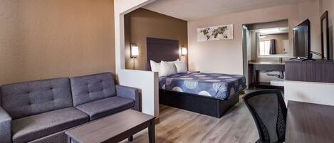 Quarto de hotel compacto com sofá, cama com colcha estampada, escrivaninha e espelho.