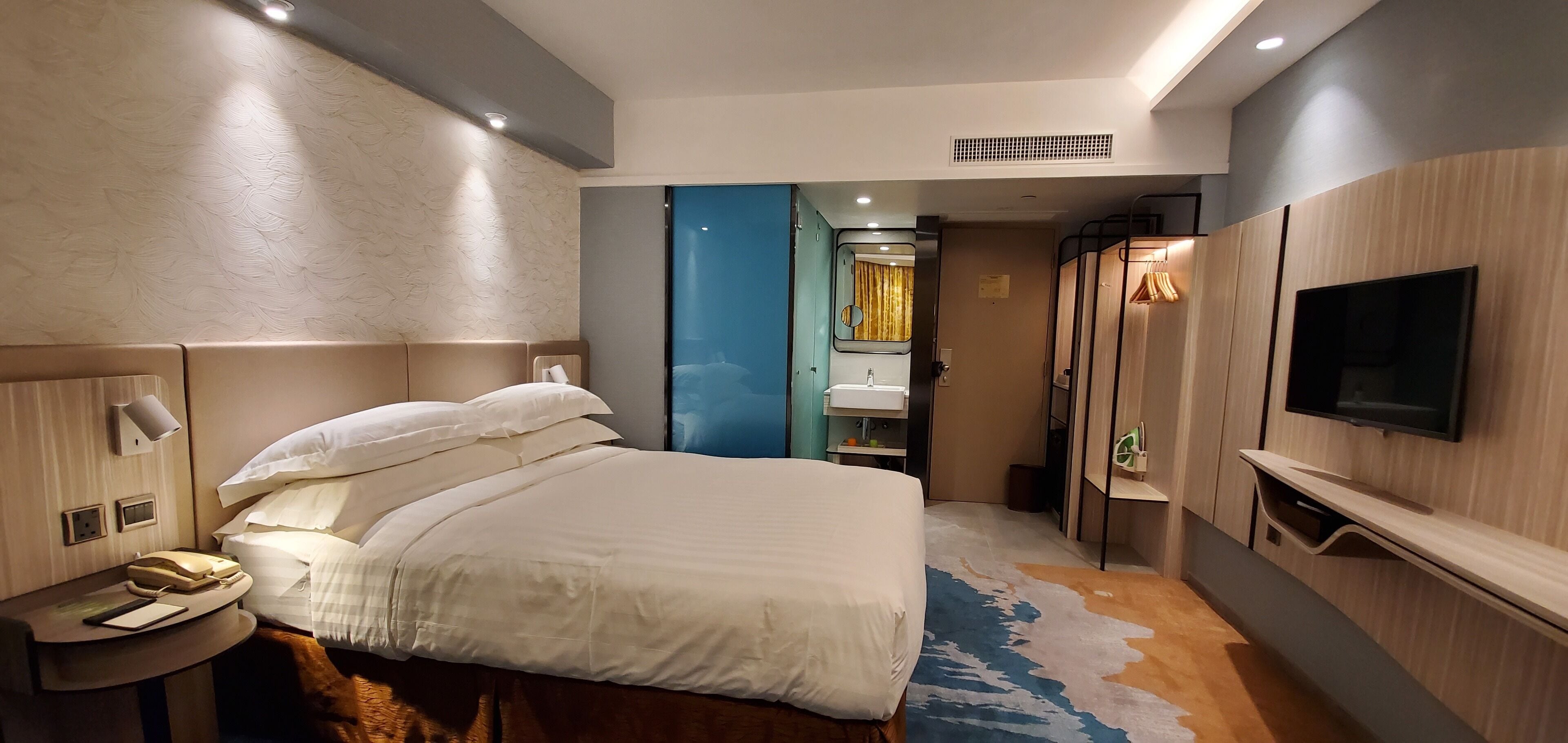 Quarto de hotel com cama, mesa de cabeceira, abajur, televisão e banheiro com espelho e porta-toalhas.