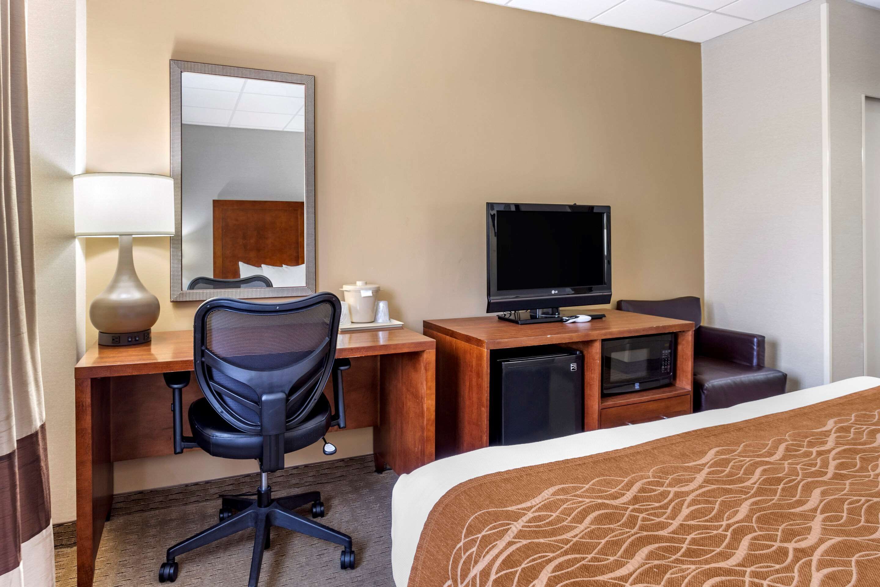 Quarto de hotel com cama, escrivaninha com computador, televisão, micro-ondas e uma cadeira.