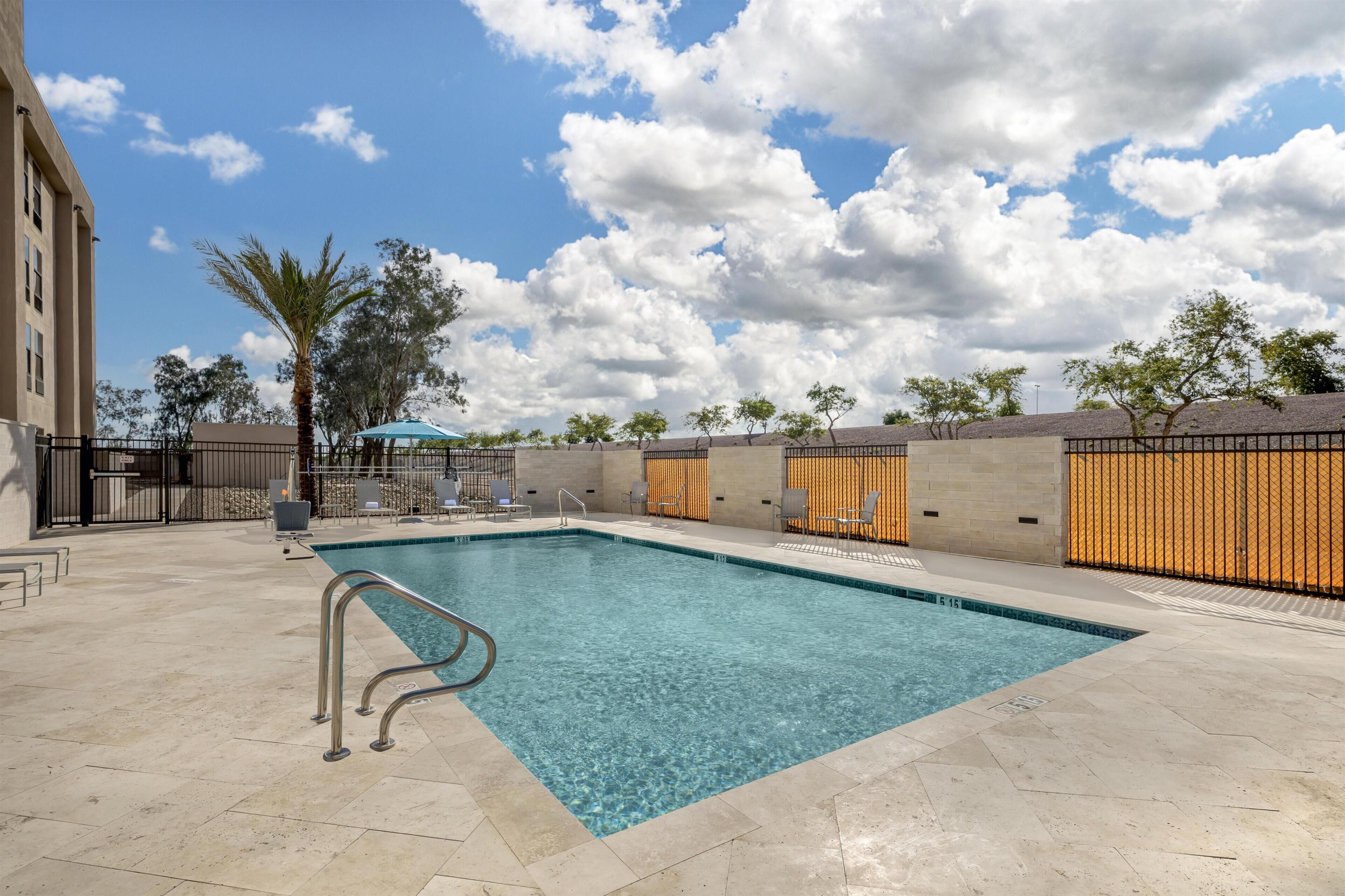 Uma piscina com escada de metal, rodeada por um deck revestido de azulejos, com uma cerca e cadeiras ao fundo.