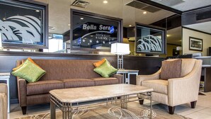 Um lobby com um sofá de couro marrom, poltrona bege e uma mesa de centro de mármore. Uma janela grande exibe o nome 'Bella Sera' e 'RESTAURANT & LOUNGE'.