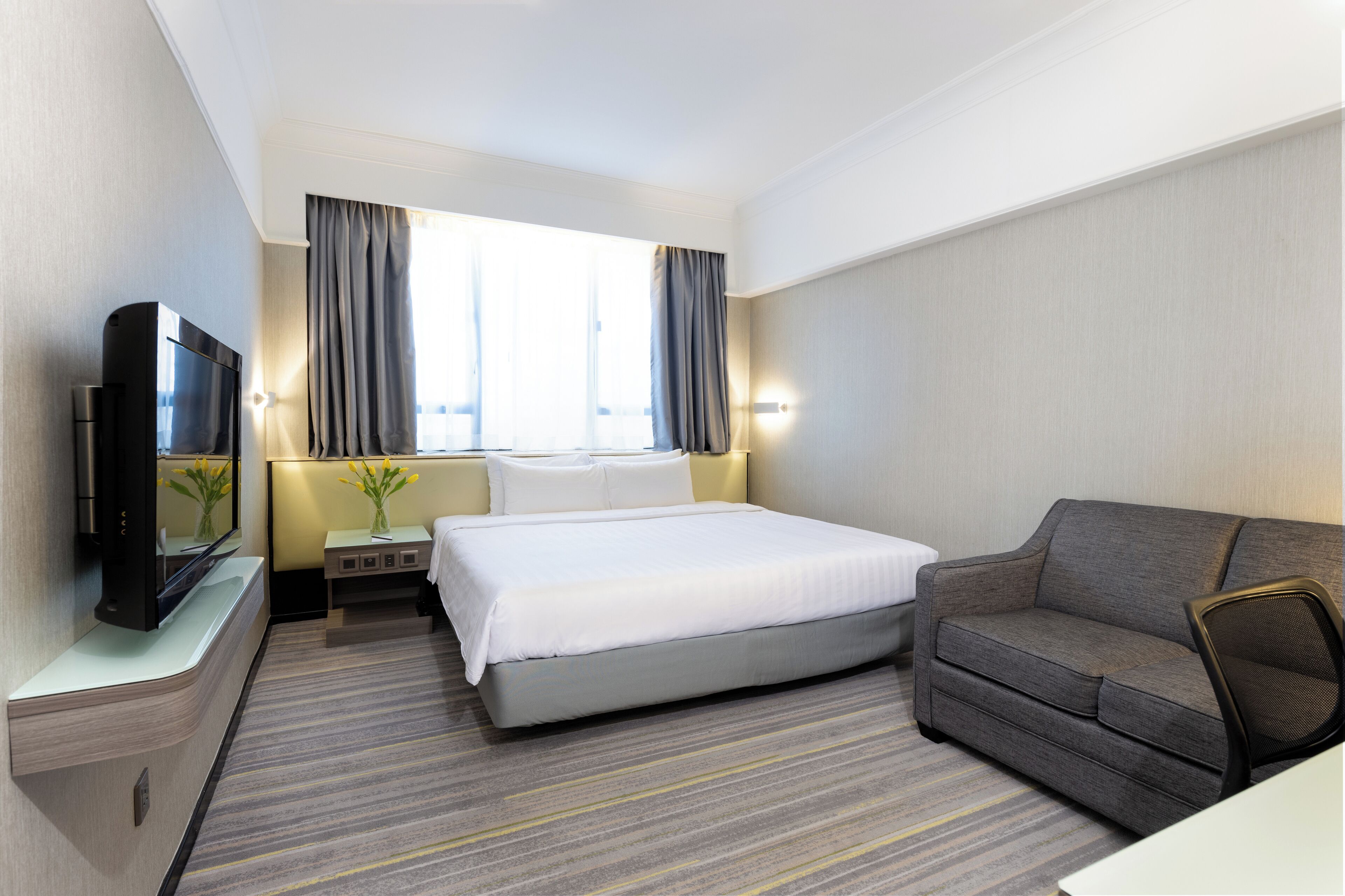 Quarto de hotel com cama, sofá, televisão, mesa de cabeceira com abajur e janela com cortinas.