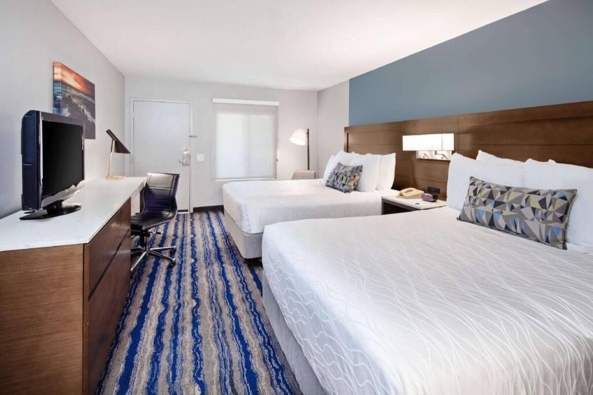 Quarto de hotel com duas camas, uma mesa de trabalho com televisão, uma cadeira e um tapete estampado.