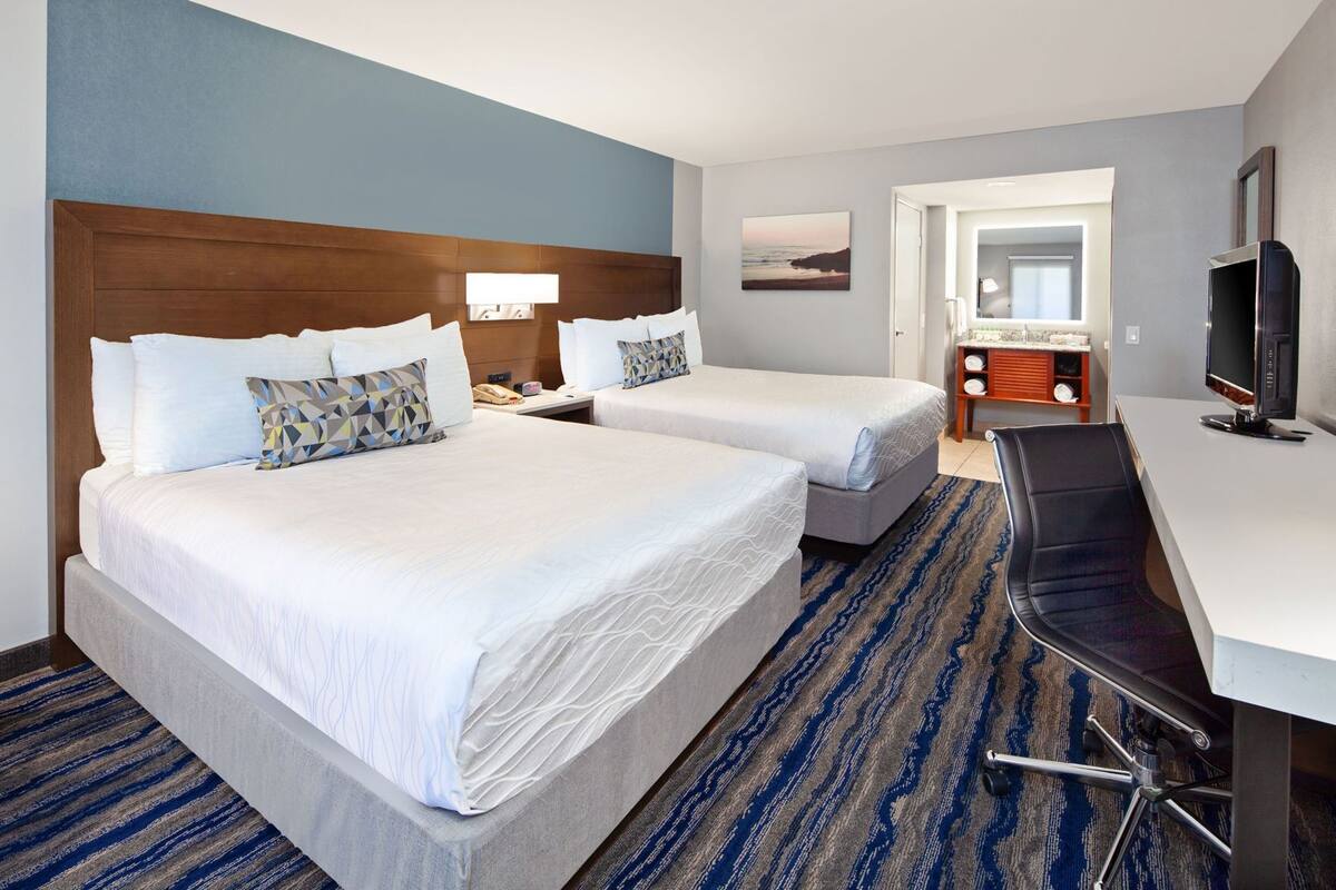 Quarto de hotel com duas camas, uma escrivaninha, uma cadeira, uma televisão e um banheiro visível através de uma porta aberta.
