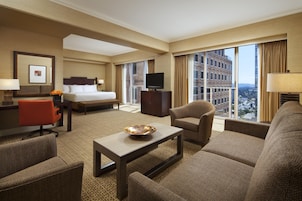 Quarto de hotel com cama, escrivaninha, televisão, sofá e vista para a paisagem urbana.