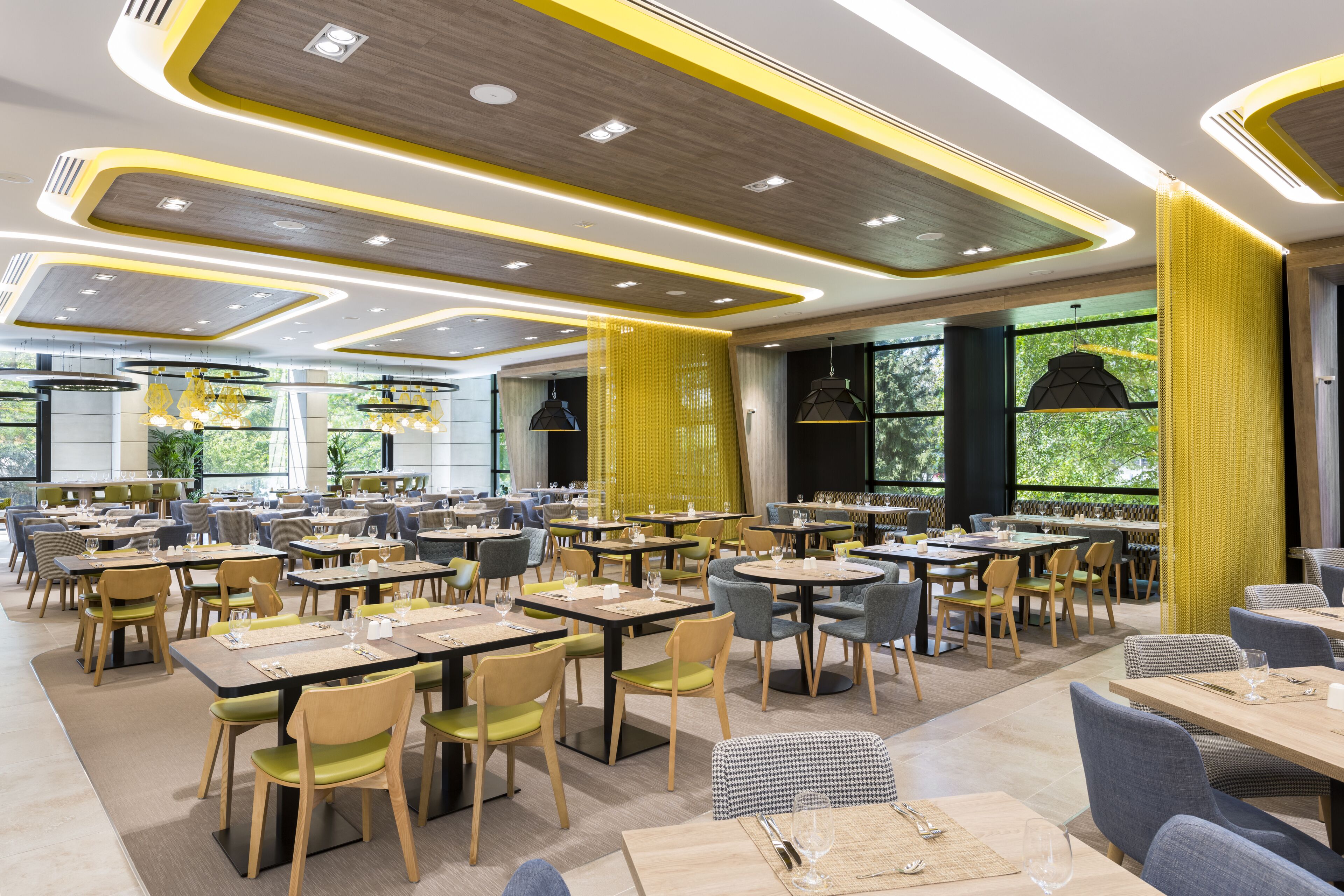 Um restaurante espaçoso com várias áreas de refeições, iluminação moderna e grandes janelas com vista para o verde.