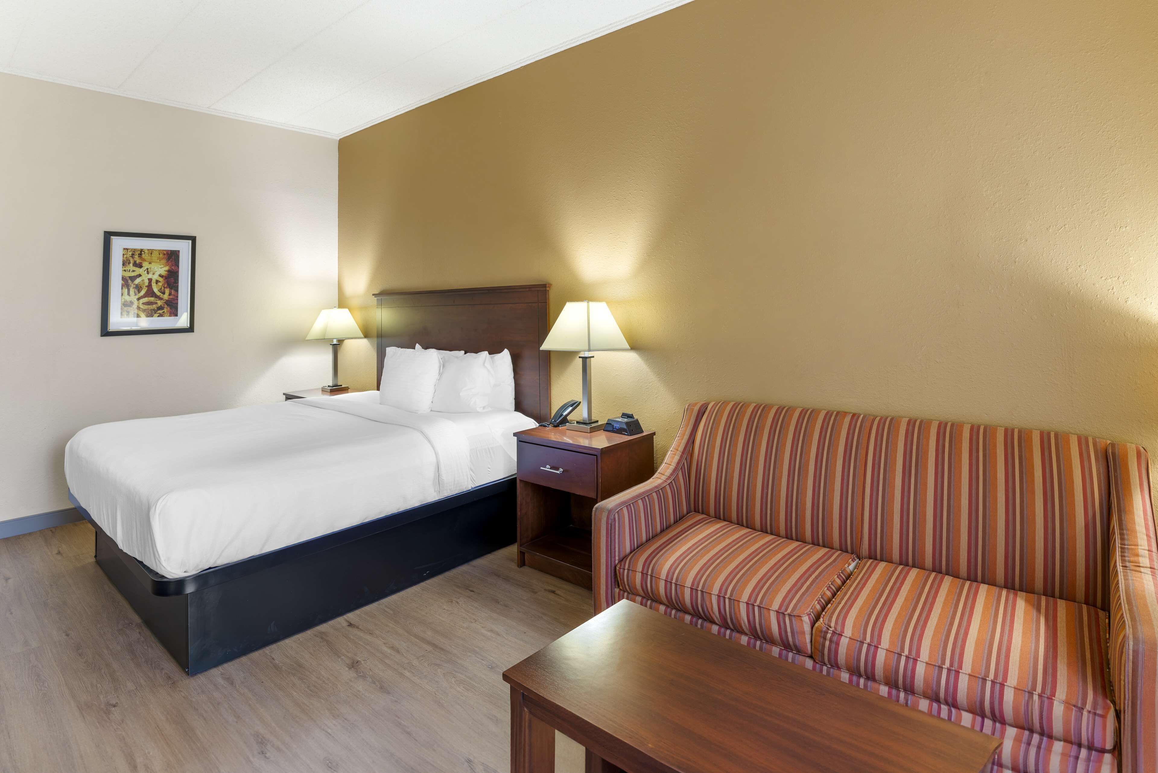 Quarto de hotel com uma cama, mesa de cabeceira, abajur e sofá listrado.