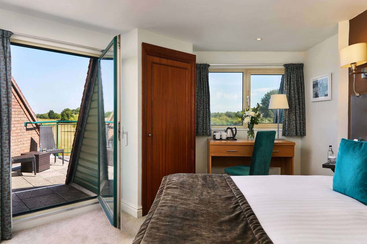 Quarto de hotel com cama, escrivaninha, cadeira, janela com cortinas e porta que dá para área externa.