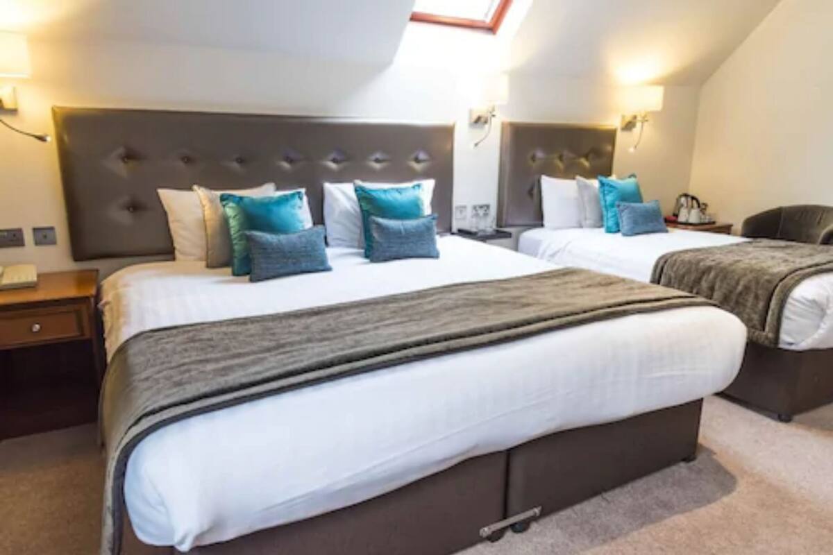 Quarto de hotel com duas camas, cada uma com cabeceira, roupa de cama branca e várias almofadas. Há uma mesa de cabeceira com abajur e telefone.