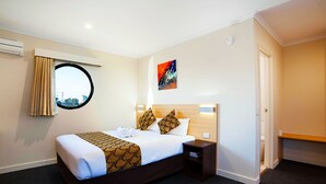 Quarto de hotel com cama, mesa de cabeceira, janela redonda e um quadro na parede.