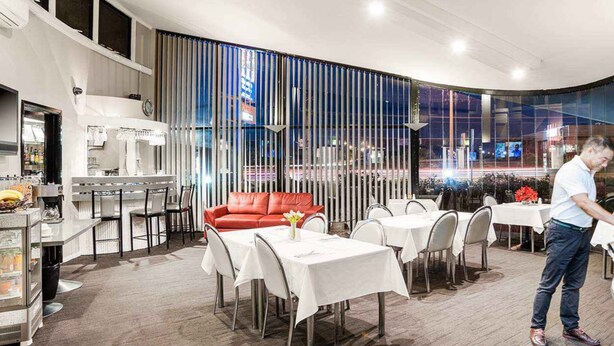 Um restaurante moderno com mesas e cadeiras brancas, um sofá vermelho e vista para a cidade à noite.