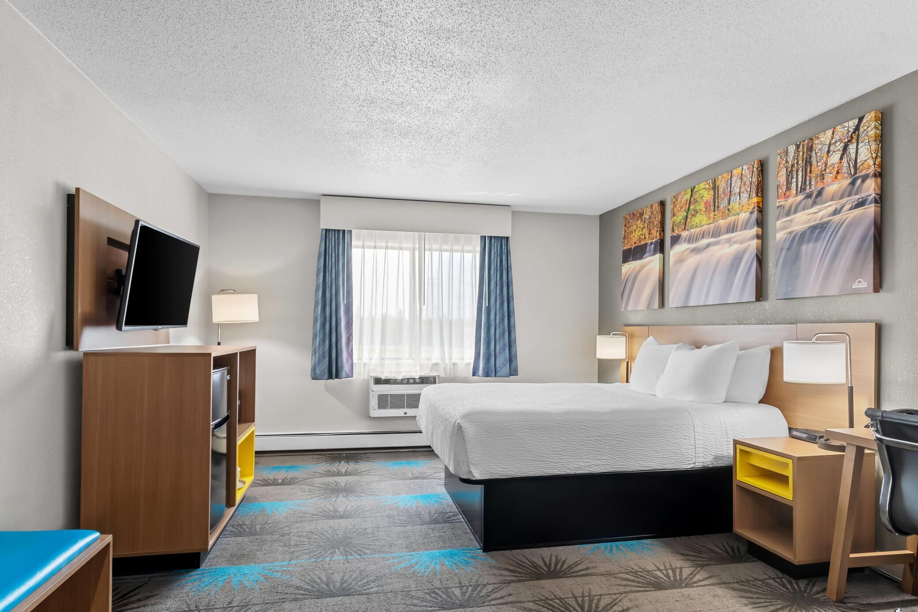Quarto de hotel com cama, secretária, televisão, janela com cortinas e obras de arte na parede.