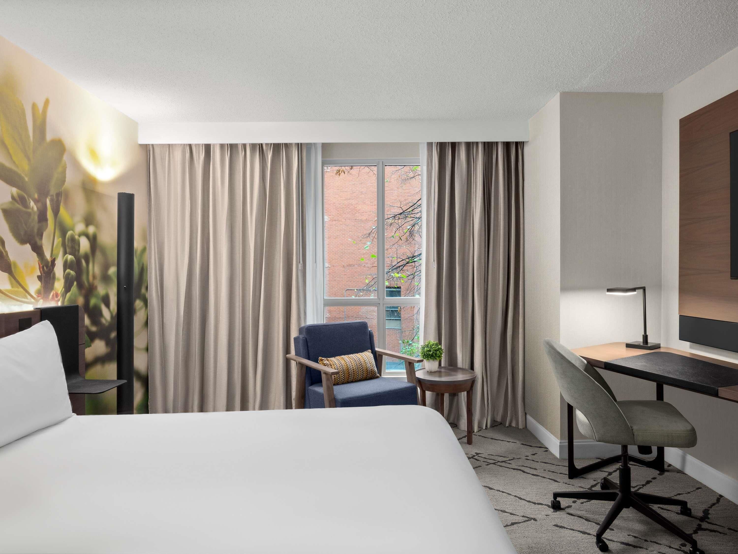 Quarto de hotel com cama, escrivaninha, cadeira e vista para um edifício de tijolos pela janela.