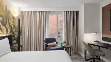 Quarto de hotel com cama, escrivaninha, cadeira e vista para um edifício de tijolos pela janela.