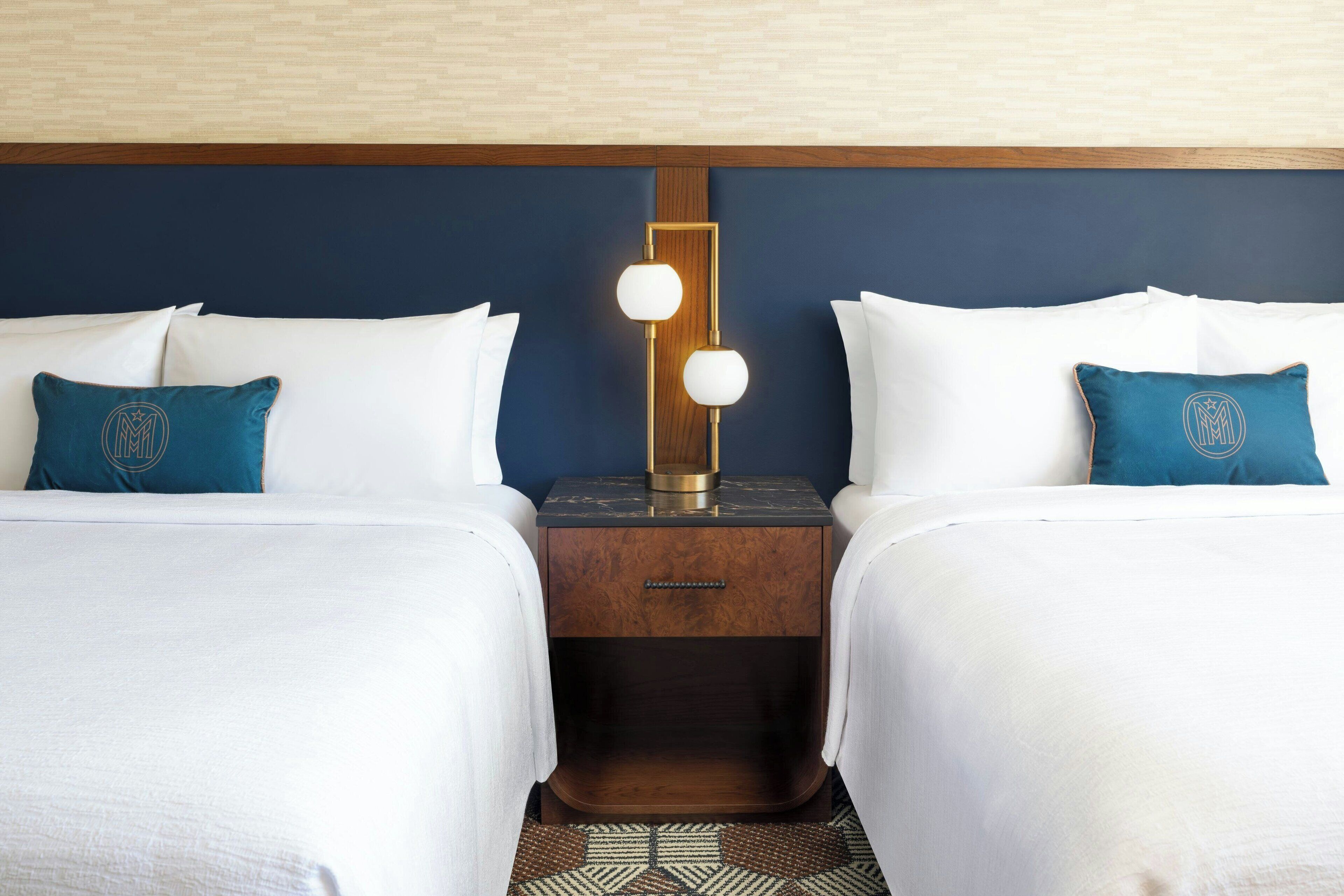 Quarto de hotel com duas camas, uma mesa de cabeceira com abajur e um tapete estampado.