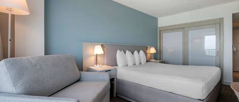 Quarto de hotel com cama, duas mesas de cabeceira com abajures, sofá e uma grande janela com vista.