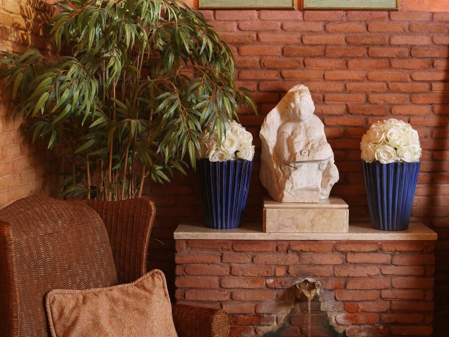 Uma cadeira de vime com almofada, uma grande planta de bambu, uma escultura de pedra e duas imagens emolduradas de abacaxis numa parede de tijolos.