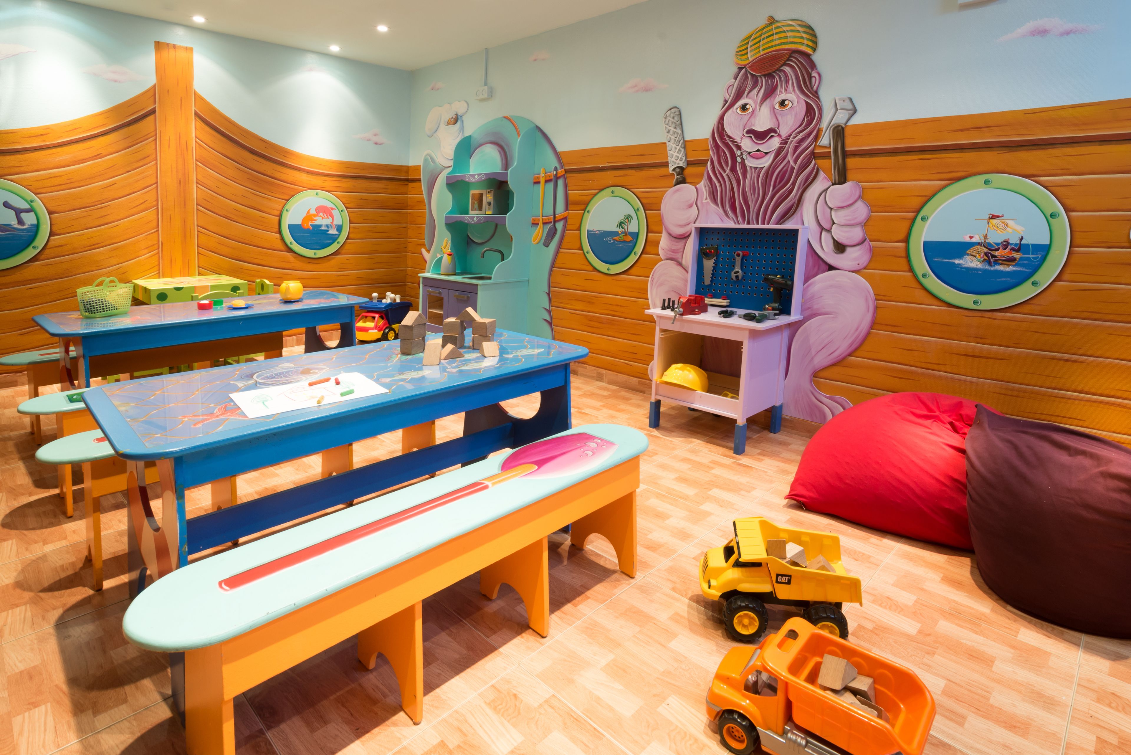 Um espaço de brincadeiras infantil com paredes de madeira, um mural grande e diversas mesas e brinquedos.
