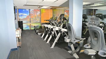 Uma academia com várias esteiras e equipamentos de exercício, uma janela grande com vista para árvores e uma televisão fixada na parede.